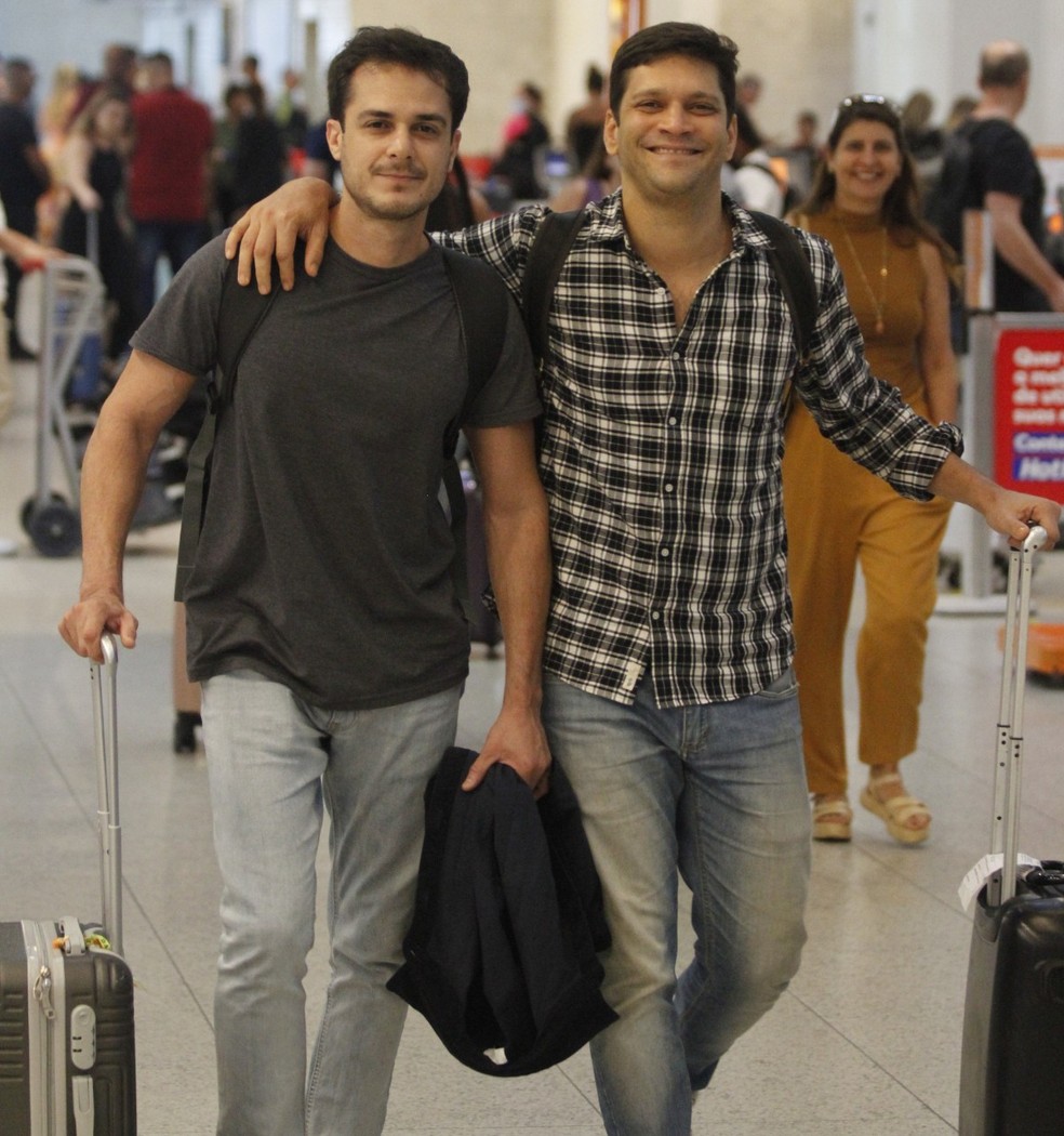 Abraçados, Armando Babaioff e o namorado desembarcam no Rio